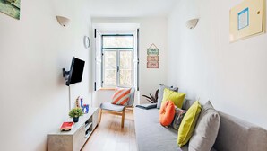Uma sala de estar com um sofá cinza, uma TV fixada na parede, uma janela com cortinas e uma pequena mesa com uma planta e livros.