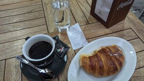 Uma mesa de madeira com uma chávena de café, um copo de água, um recibo e um croissant num prato.