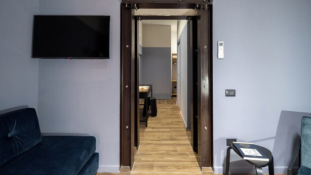 Um corredor com porta de madeira escura, TV de tela plana na parede, sofá azul e uma pequena mesa com um livro.