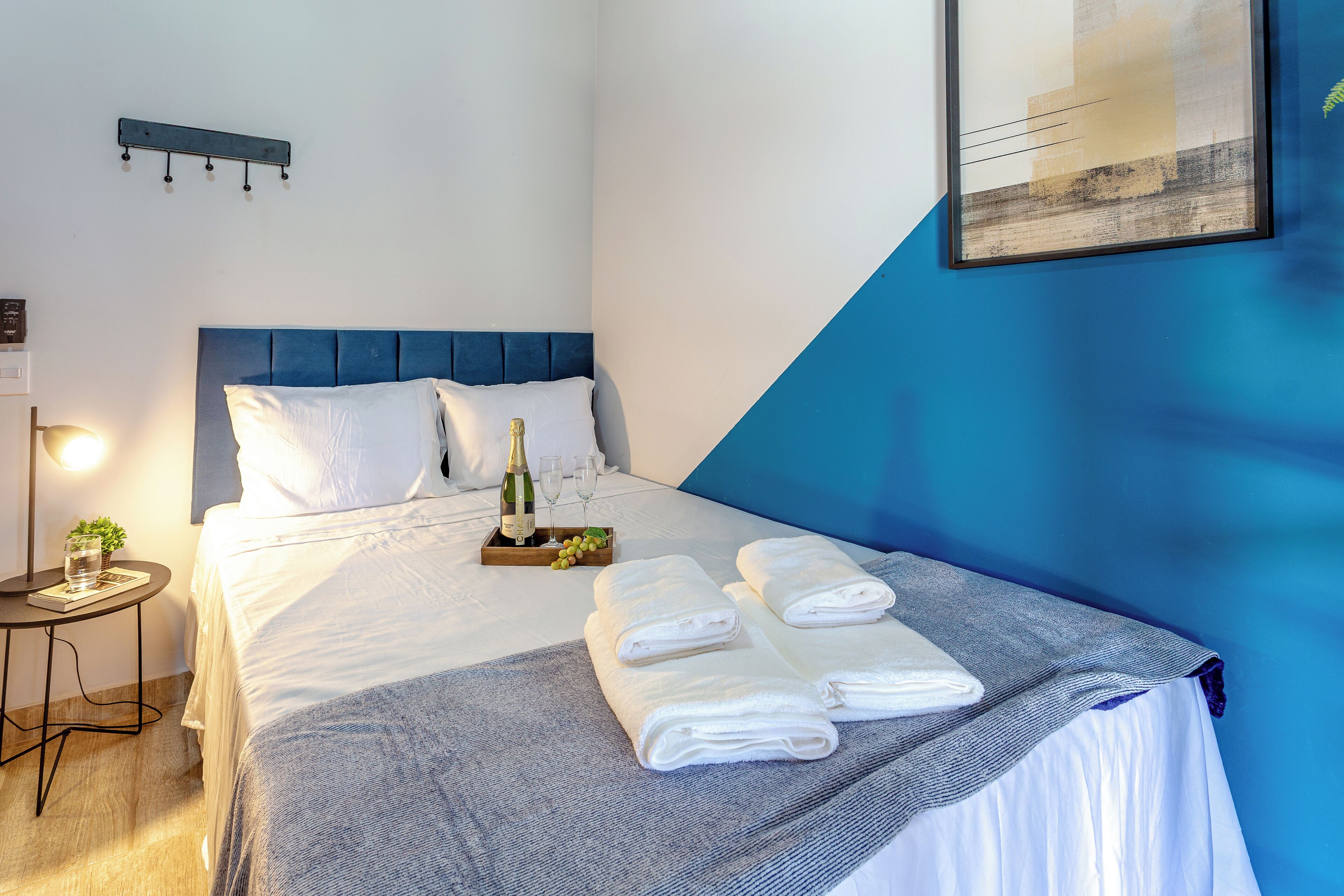 Cama bem arrumada com lençóis brancos, cabeceira azul e mesa de cabeceira com abajur e uma pequena planta.