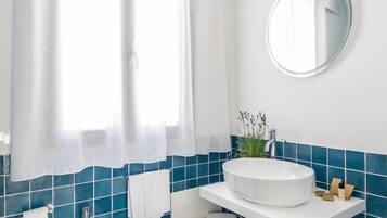 Casa de banho com lavatório branco, azulejos azuis, espelho e janela com cortinas de tule.