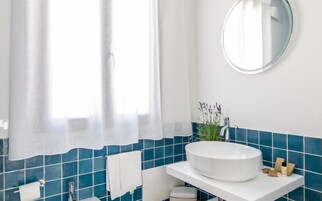 Casa de banho com lavatório branco, azulejos azuis, espelho e janela com cortinas de tule.