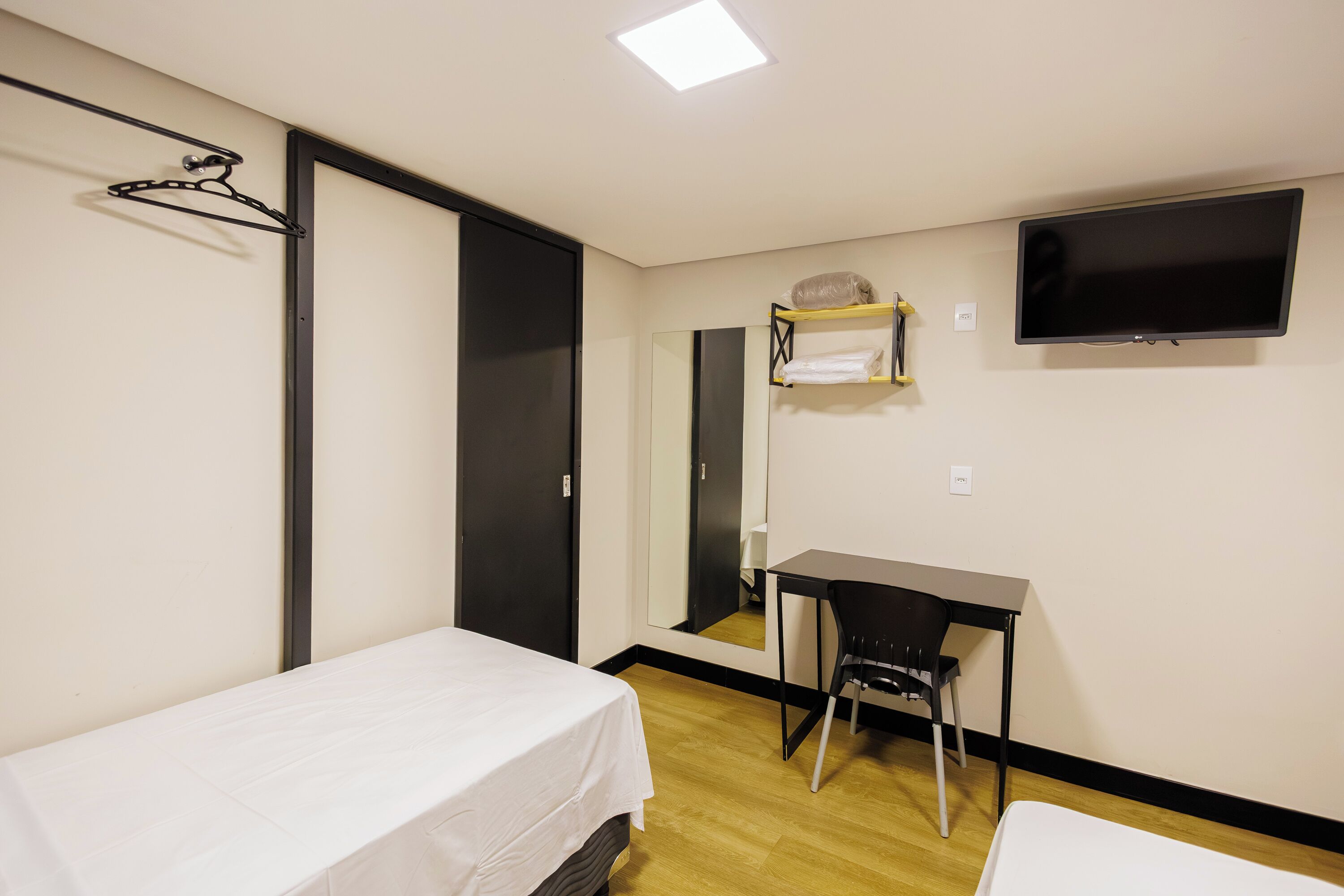 Quarto de hotel com cama, escrivaninha, cadeira, televisão e espelho.