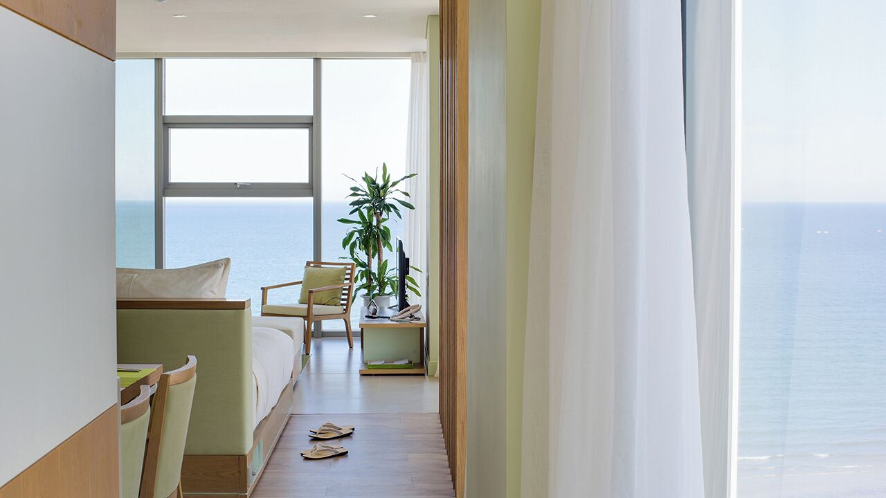 Quarto de hotel com cama, escrivaninha, cadeira e vista para o mar.