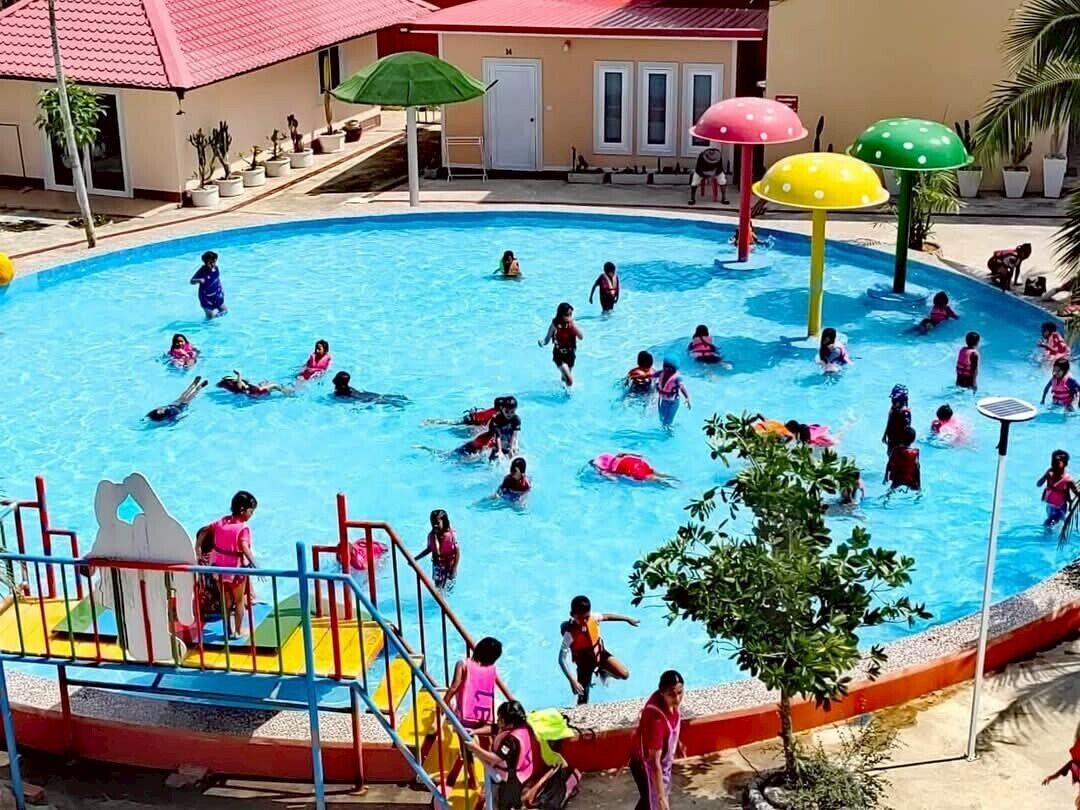Piscina com várias pessoas, rodeada por um edifício com telhado vermelho e plantas em vasos.