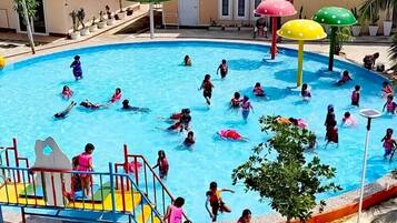 Piscina com várias pessoas, rodeada por um edifício com telhado vermelho e plantas em vasos.