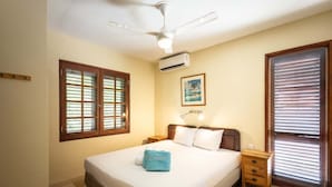 Quarto bem arrumado com uma cama, mesa de cabeceira, um abajur, ventilador de teto, ar condicionado e janela com persianas.