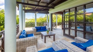 Um pátio externo com mobiliário de vime, uma mesa e grandes janelas com vista para o verde.