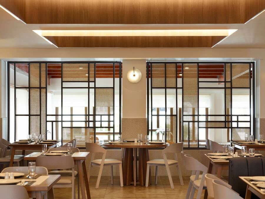 Um restaurante moderno com mesas e cadeiras de madeira, grandes janelas com painéis decorativos e uma luminária de teto aconchegante.