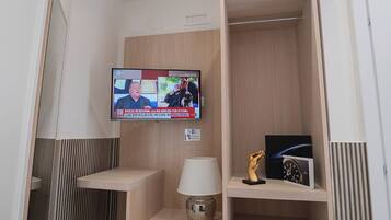 Quarto de hotel moderno com televisão de tela plana na parede, mesa de cabeceira com abajur, prateleira pequena com objeto decorativo e armários embutidos.