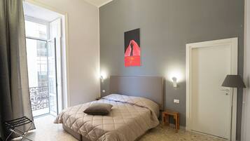 Quarto com uma cama, mesa de cabeceira pequena, abajur, porta e janela com vista para edifícios.