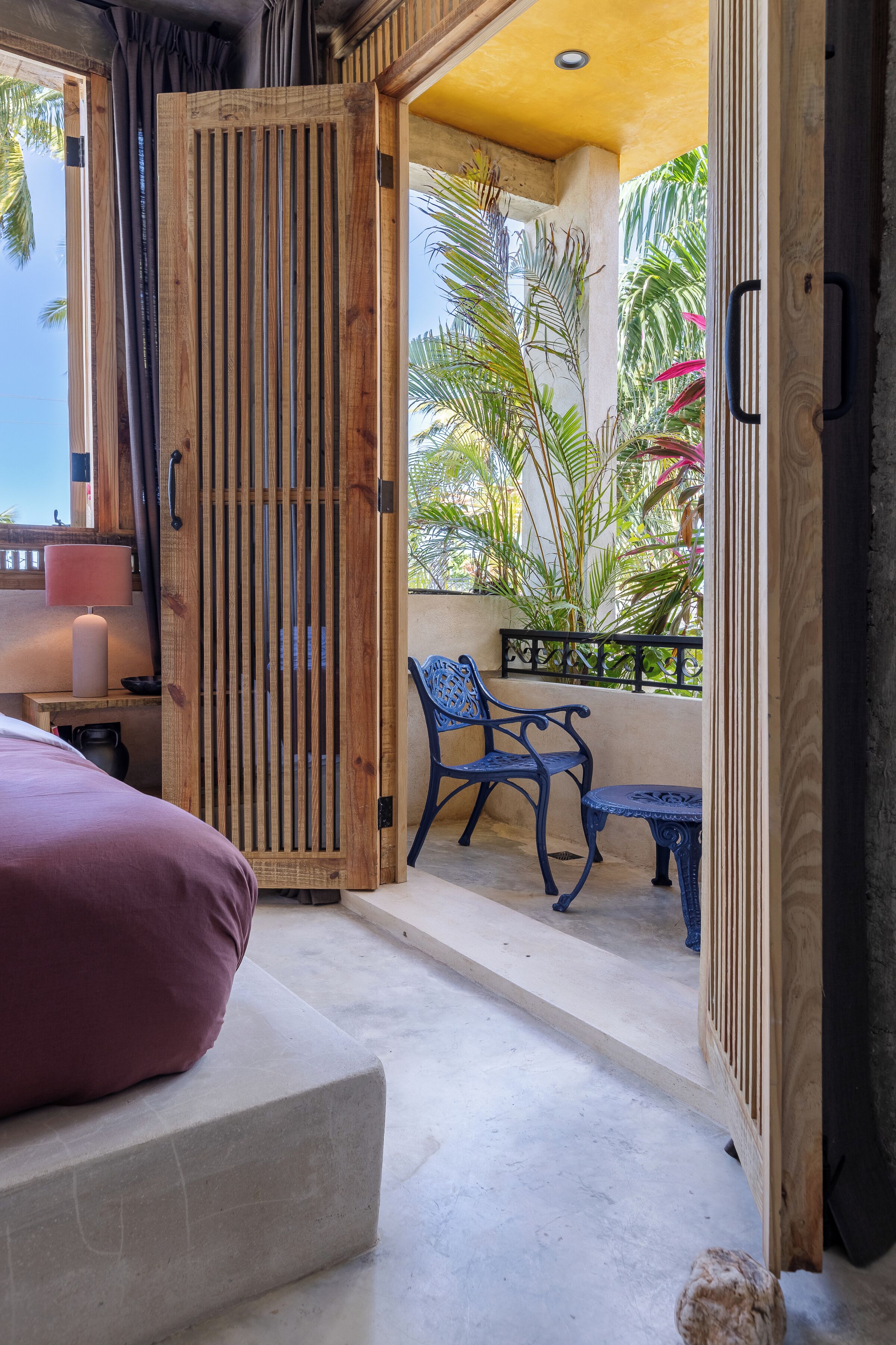 Quarto com cama, porta de madeira que dá para área externa com mesa, cadeiras e plantas.