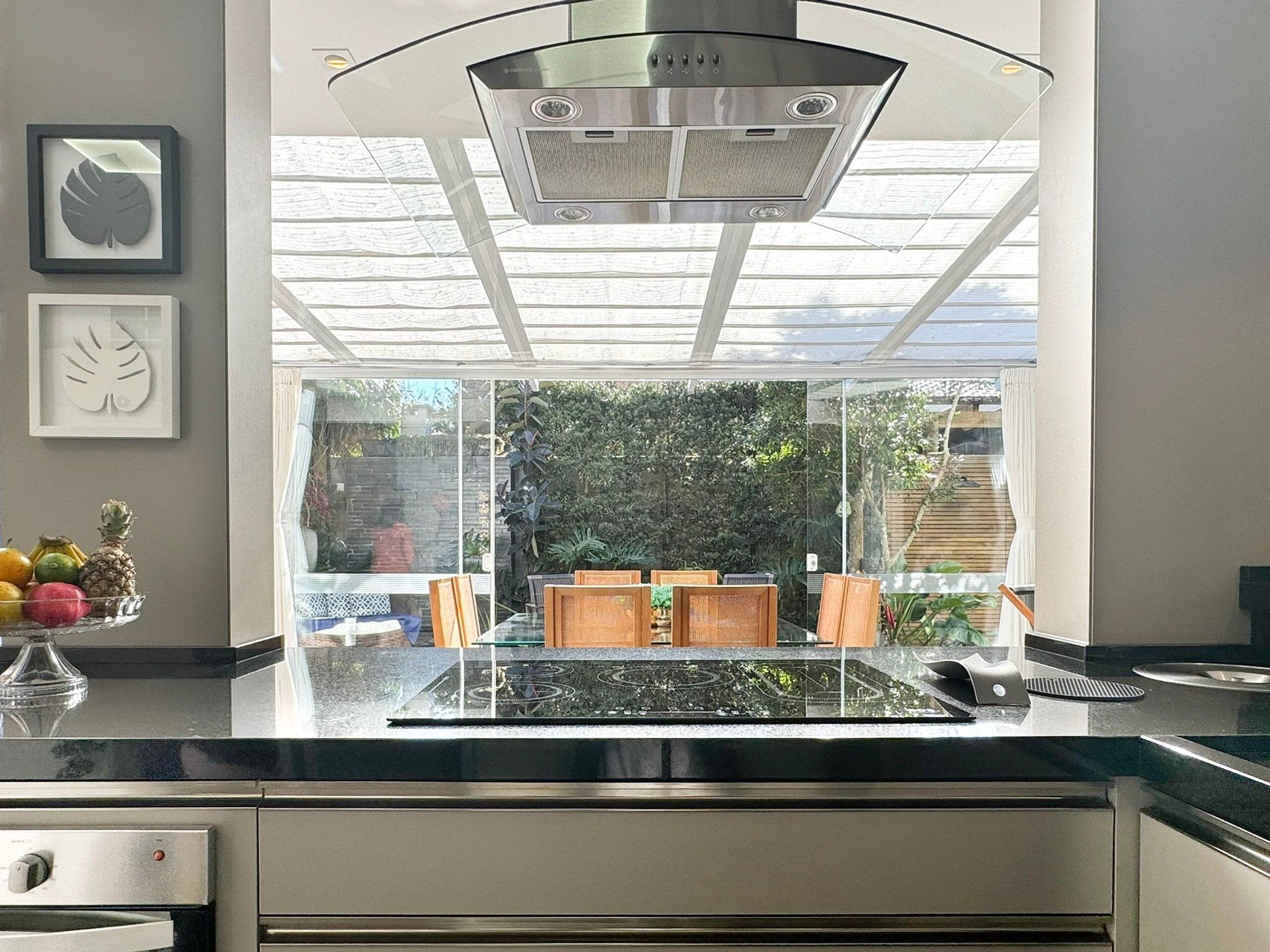 Cozinha moderna com divisória de vidro, exaustor de aço inoxidável e vista para um jardim através do vidro.