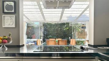 Cozinha moderna com divisória de vidro, exaustor de aço inoxidável e vista para um jardim através do vidro.