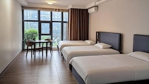 Quarto de hotel com três camas, uma pequena mesa com uma cadeira e uma janela grande com cortinas.