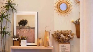 Um quarto com uma obra de arte emoldurada, uma mesa consola de madeira com objetos decorativos, um espelho embutido em formato de sol e uma placa com texto.