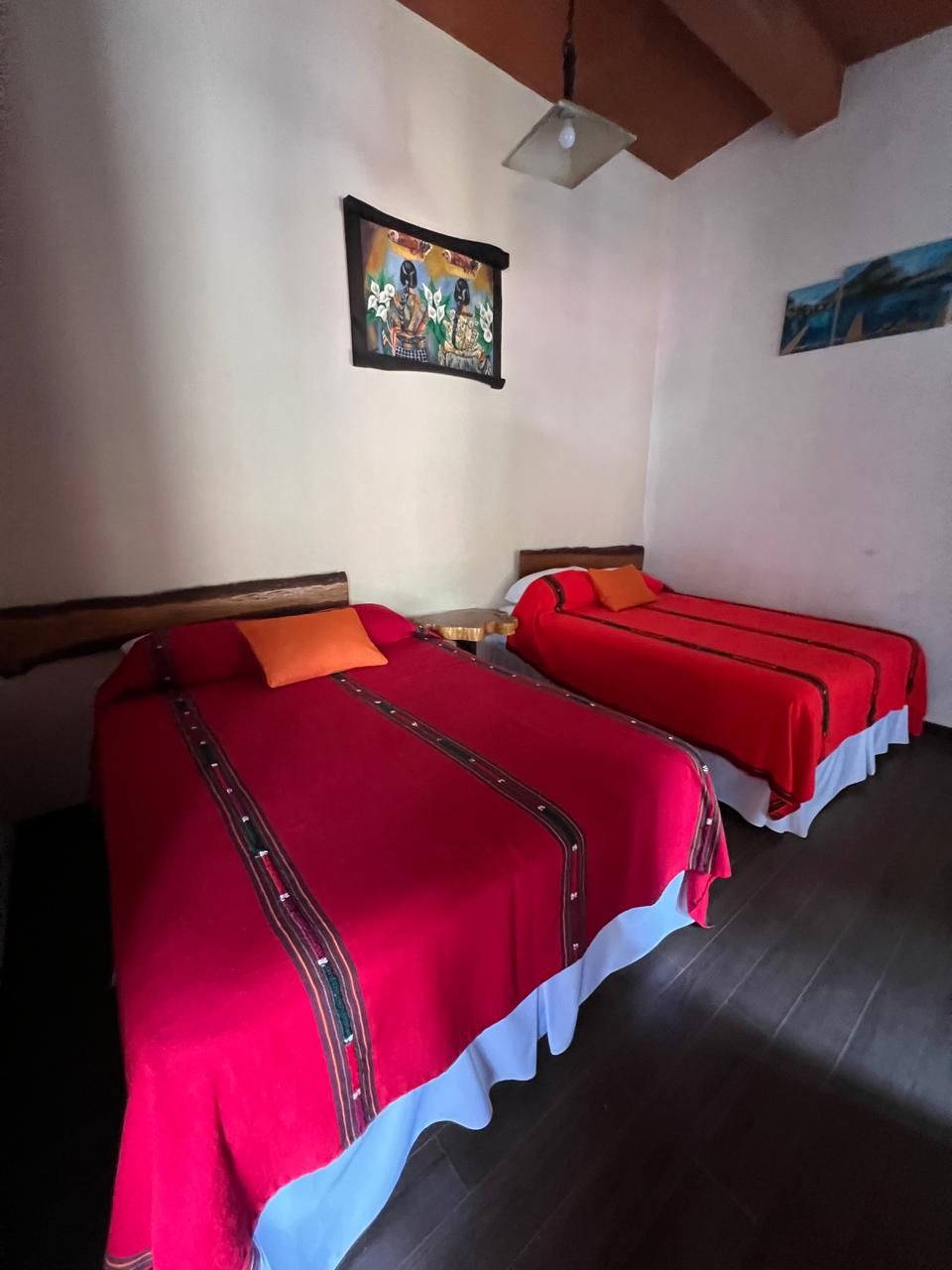 Quarto com duas camas, cada uma com roupa de cama vermelha e acabamento branco. Há um ventilador de teto e uma pintura na parede.
