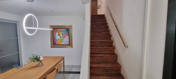 Uma escadaria com degraus de madeira e corrimão, uma área de jantar com mesa e cadeiras de madeira, e uma pintura abstrata emoldurada na parede.