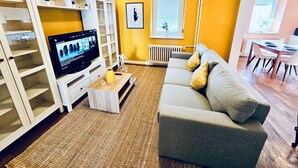 Sala de estar com sofá cinza, televisão em suporte de madeira, armário branco com portas de vidro e janela com persianas.