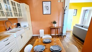 Uma sala de jantar com uma mesa de madeira posta para quatro, cadeiras brancas e uma cozinha com armários brancos e uma pia.