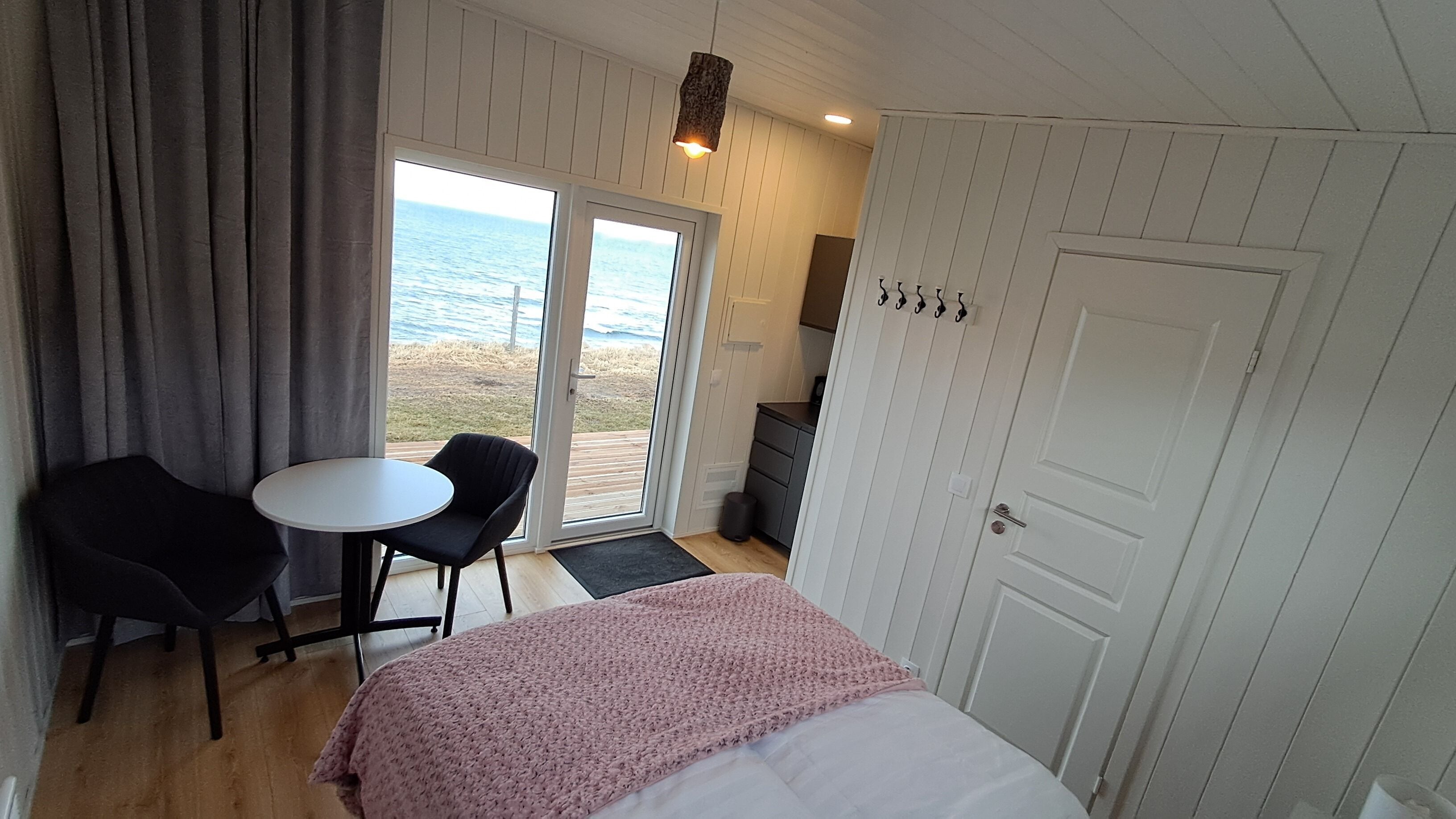 Quarto com cama, mesinha, duas cadeiras e vista para o mar através de uma porta de correr, além de uma porta branca com ganchos.