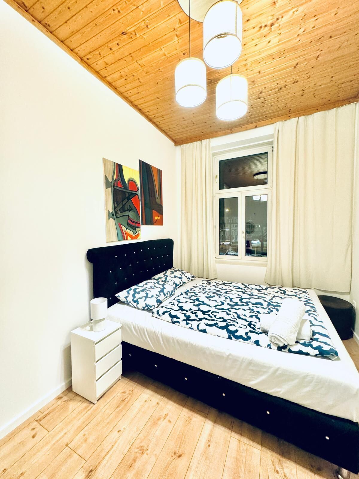 Quarto com teto de madeira, cama com roupa de cama estampada, mesa de cabeceira e janela com cortinas.