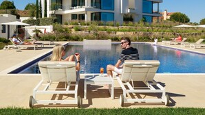 Uma área moderna à beira da piscina, com espreguiçadeiras e vista desobstruída para um edifício de vários andares ao fundo.