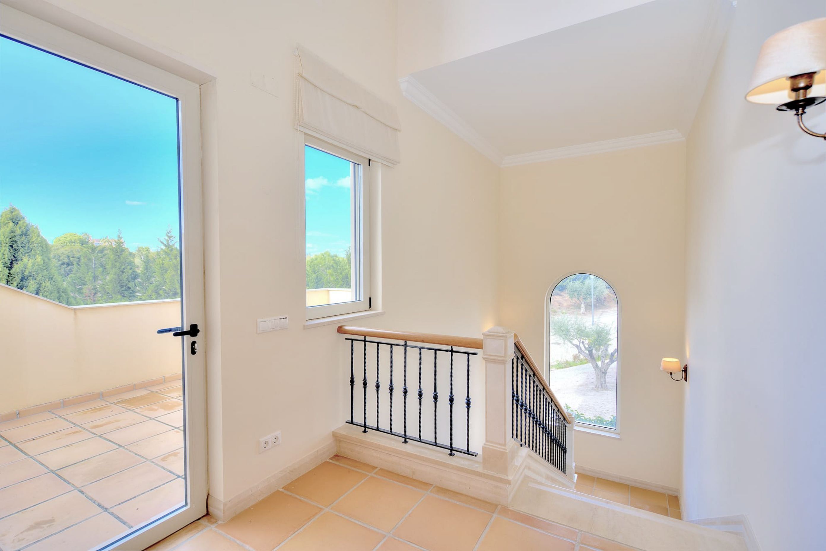 Uma varanda ampla com vista clara para árvores e céu, uma escadaria com corrimão e uma porta que dá acesso a uma área externa.