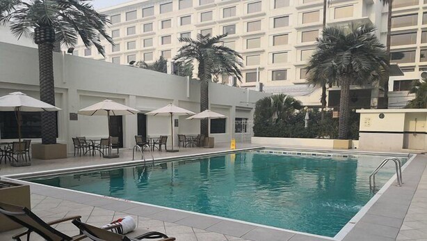 Piscina com espreguiçadeiras e guarda-sóis, cercada por palmeiras e um edifício ao fundo.