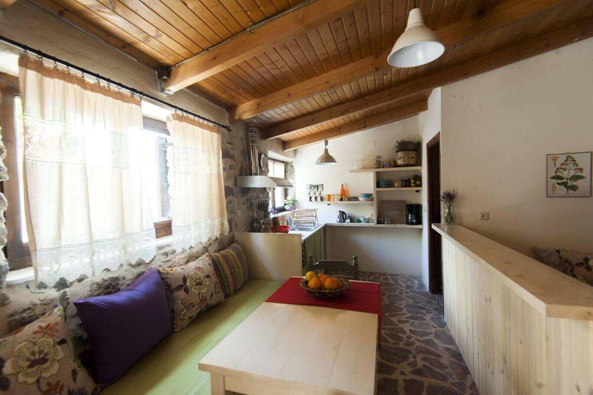 Interior acolhedor e rústico, com teto de madeira, área de jantar com mesa e cadeiras, e cozinha ao fundo.