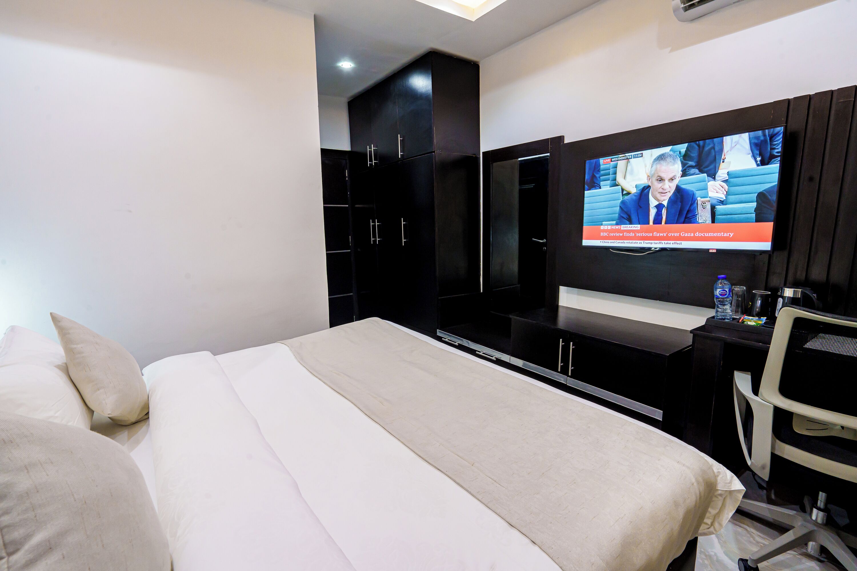 Quarto de hotel com cama, televisão na parede, escrivaninha com cadeira e guarda-roupa.