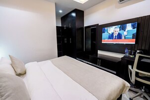 Quarto de hotel com cama, televisão na parede, escrivaninha com cadeira e guarda-roupa.