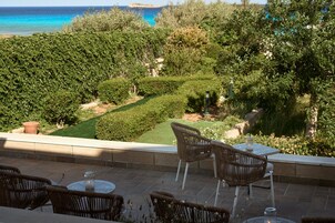 Área externa com mesas e cadeiras de vime, com vista para um jardim bem cuidado e o mar.