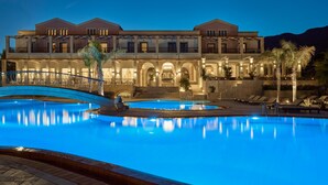Um hotel grande, com vários andares, piscina, área externa com mesas e uma entrada bem iluminada.