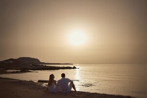 Um casal sentado na praia ao pôr do sol, com o mar calmo e um afloramento rochoso ao longe.
