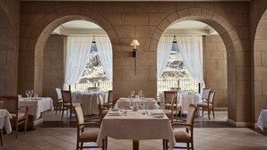 Um restaurante com uma mesa comprida posta para refeições, paredes de pedra arqueadas e cortinas sobre as janelas.