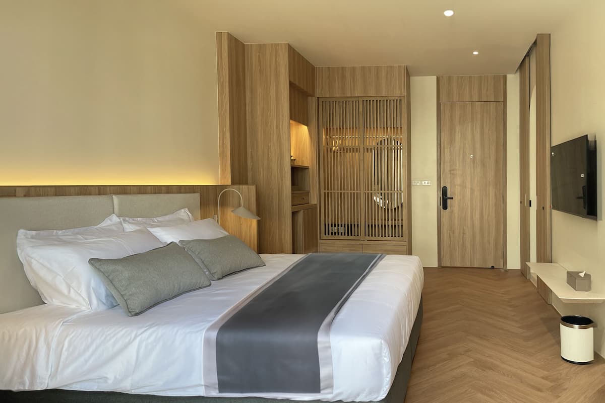 Quarto de hotel moderno com uma cama grande, cabeceira de madeira, mesa de cabeceira com abajur, televisão na parede e guarda-roupa de madeira.