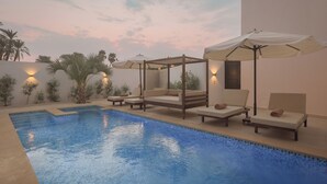 Piscina com espreguiçadeiras e guarda-sóis, cercada por plantas e uma parede branca.