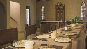 Uma sala de jantar com uma mesa comprida posta para refeição, rodeada por cadeiras, com uma escada à esquerda e uma janela com vista para o verde.