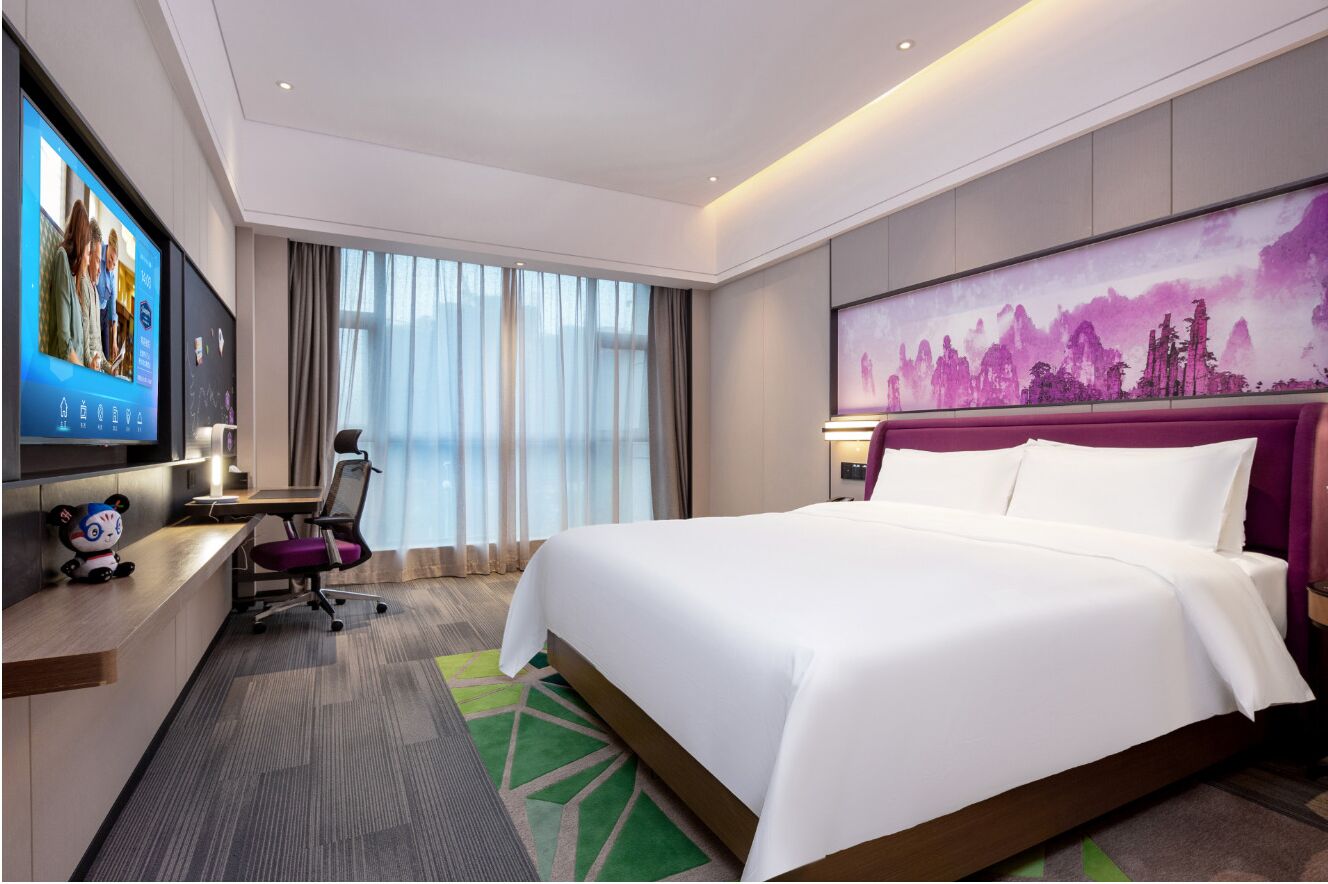 Quarto de hotel com uma cama grande, uma escrivaninha com computador, televisão e um quadro na parede.