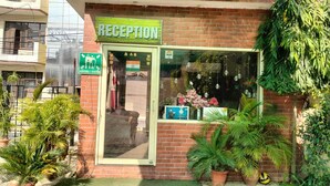 Uma área de recepção com dossel verde, uma placa indicando um ponto de encontro e plantas em vasos ao redor da entrada.