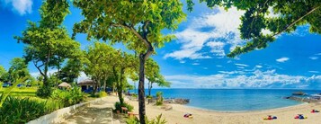 Uma praia com uma passarela bordeada por árvores, um guarda-sol e uma pequena construção ao fundo.