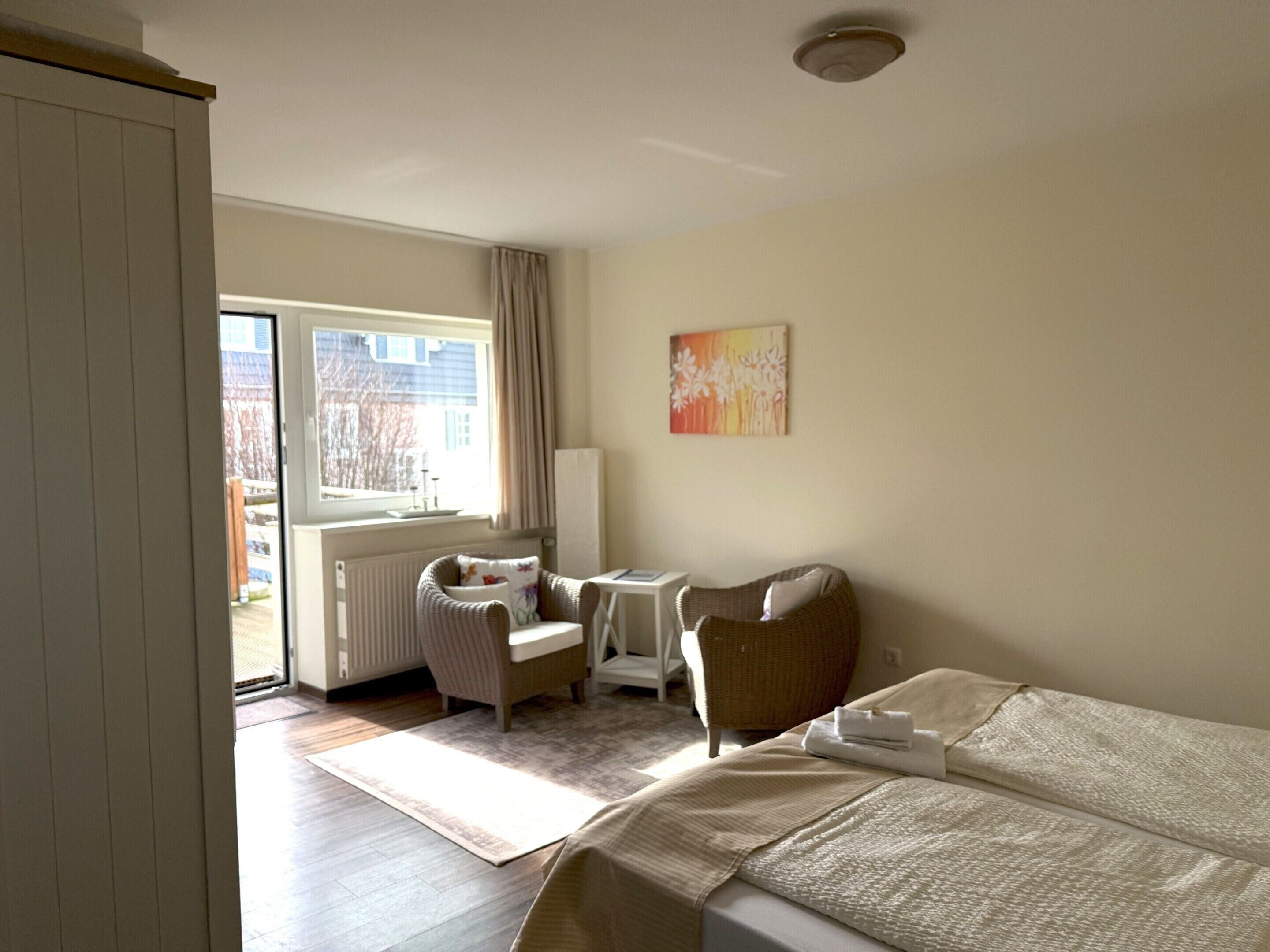 Quarto de hotel com cama, duas cadeiras, uma mesinha, radiador e janela com cortinas.