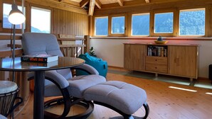 Interior acolhedor de uma cabana com teto de madeira, mesa de jantar com livros, uma poltrona estofada, um banco e um armário com janelas que dão para uma área arborizada.