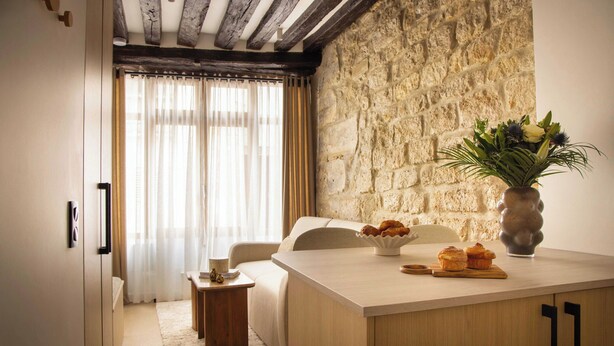 Um quarto aconchegante com parede de pedra, teto de madeira, uma cama, uma mesinha com doces e um vaso com flores.