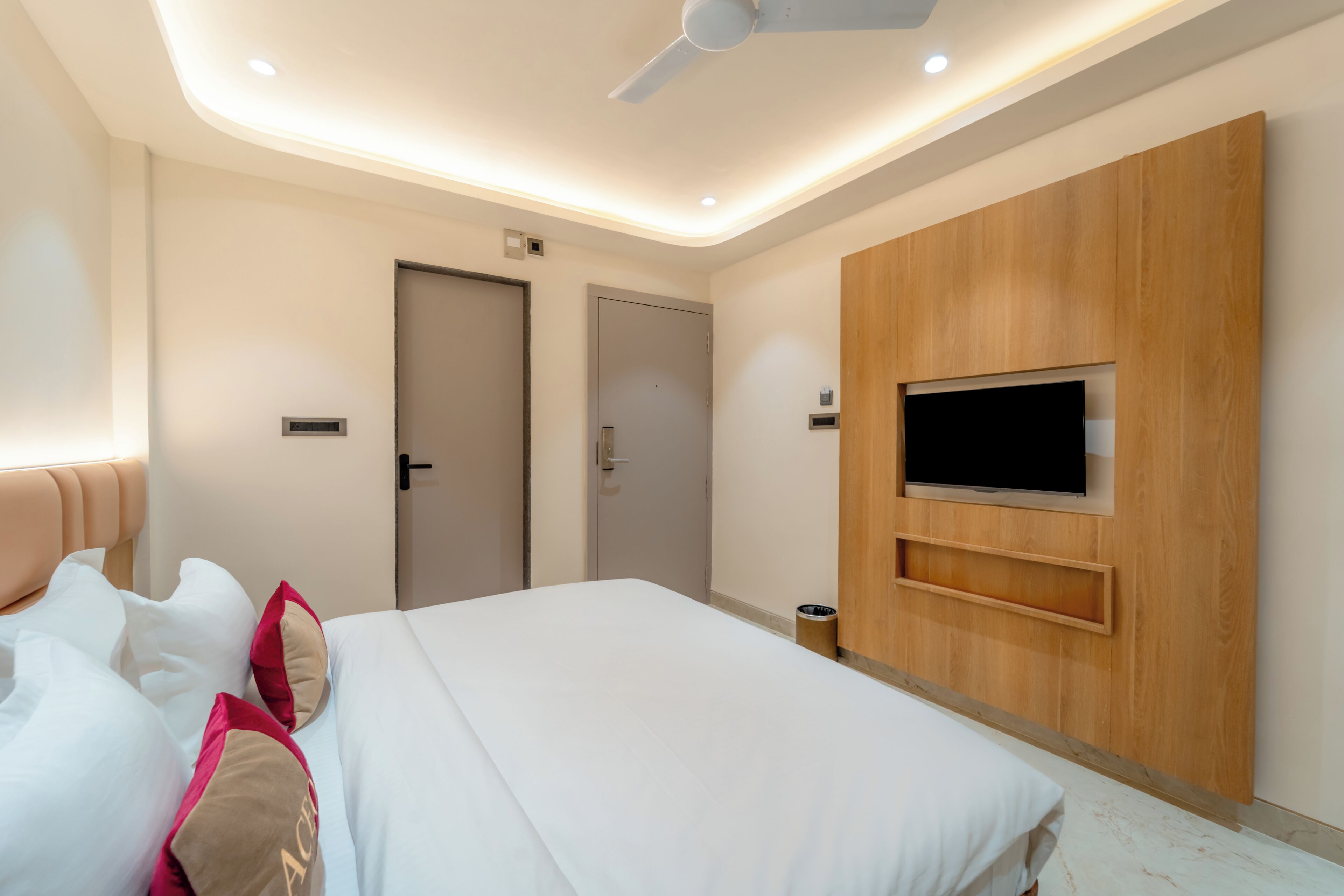 Quarto de hotel moderno com uma cama grande, um ventilador de teto, uma TV fixada em um painel de madeira e uma porta.