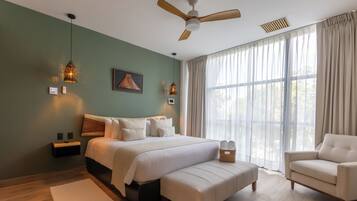 Quarto com cama, mesa de cabeceira, banco, cadeira, ventoinha no teto e grandes janelas com cortinas.