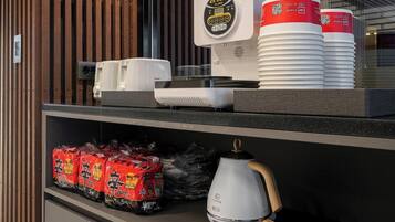 Uma área de cozinha com cafeteira, xícaras brancas empilhadas e alimentos embalados nas prateleiras.
