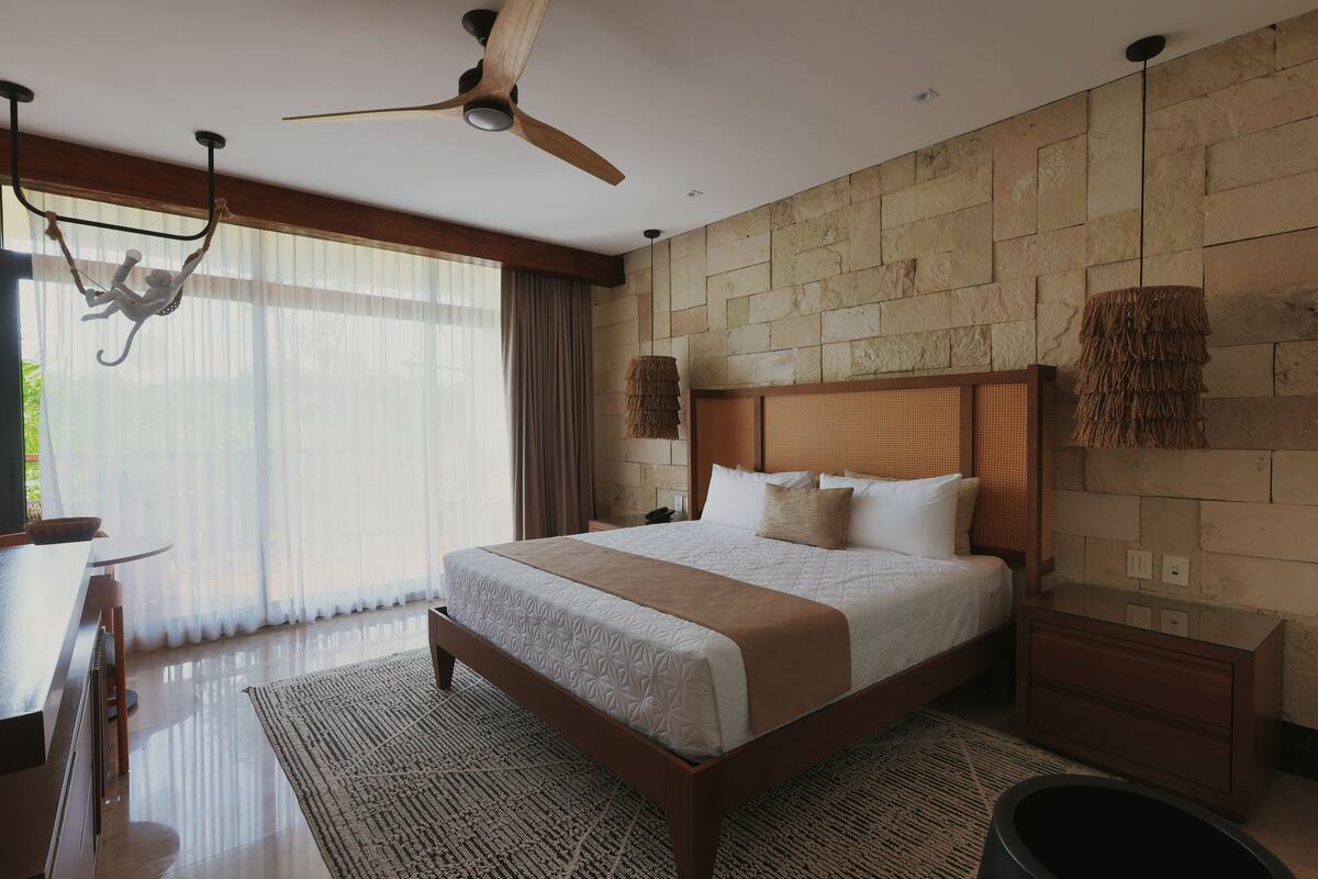 Quarto de hotel moderno com uma cama grande, um ventilador de teto, parede de pedra e vista para o exterior.
