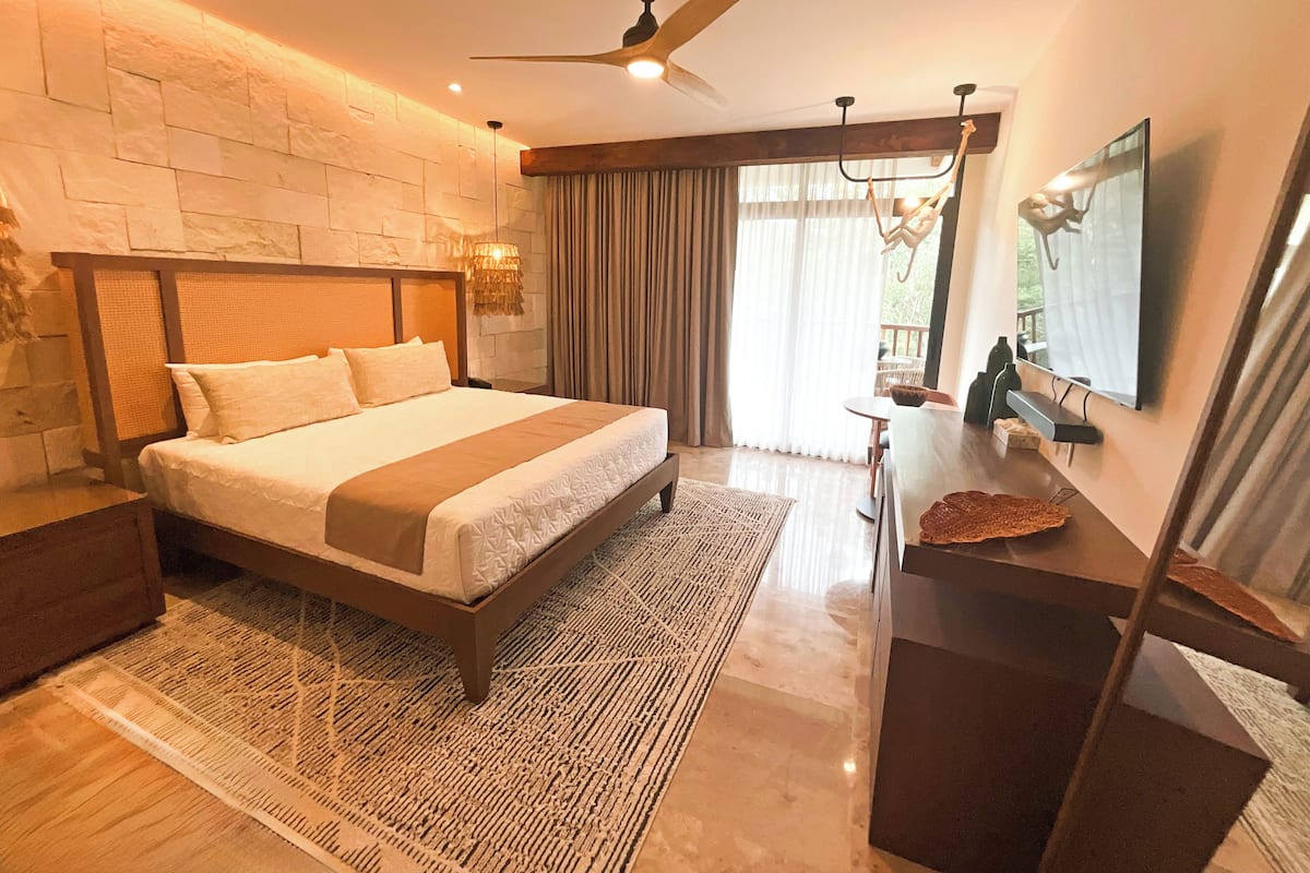 Quarto moderno com cama grande, ventilador de teto, parede de pedra e vista para o exterior.
